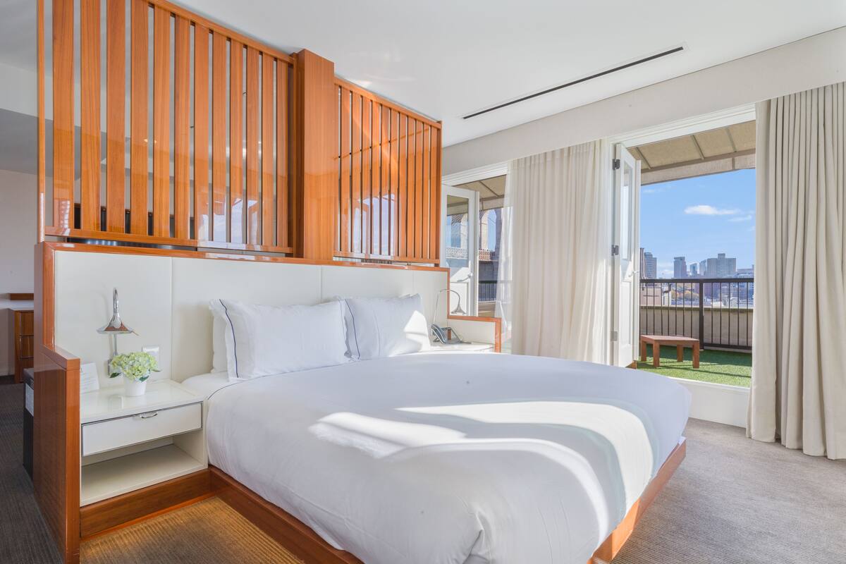 Quarto de hotel moderno com uma cama grande, cabeceira de madeira, uma mesa de cabeceira com abajur e vista da cidade através de amplas janelas.