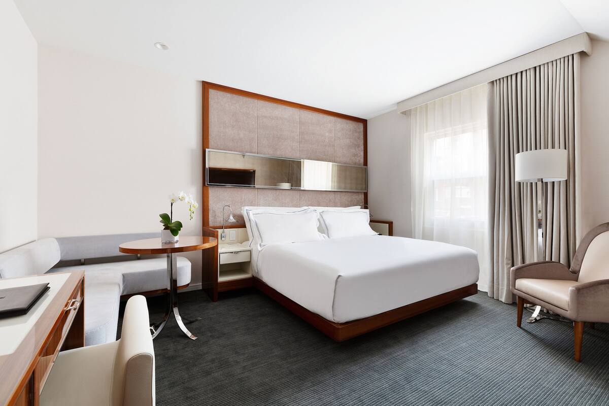 Quarto de hotel com cama, escrivaninha, cadeira, mesinha com planta e luminária.