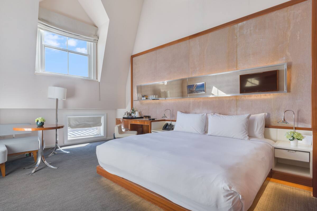 Quarto de hotel moderno com uma cama grande, uma escrivaninha, uma cadeira, uma mesinha, uma luminária, um espelho e uma janela com vista para o céu.