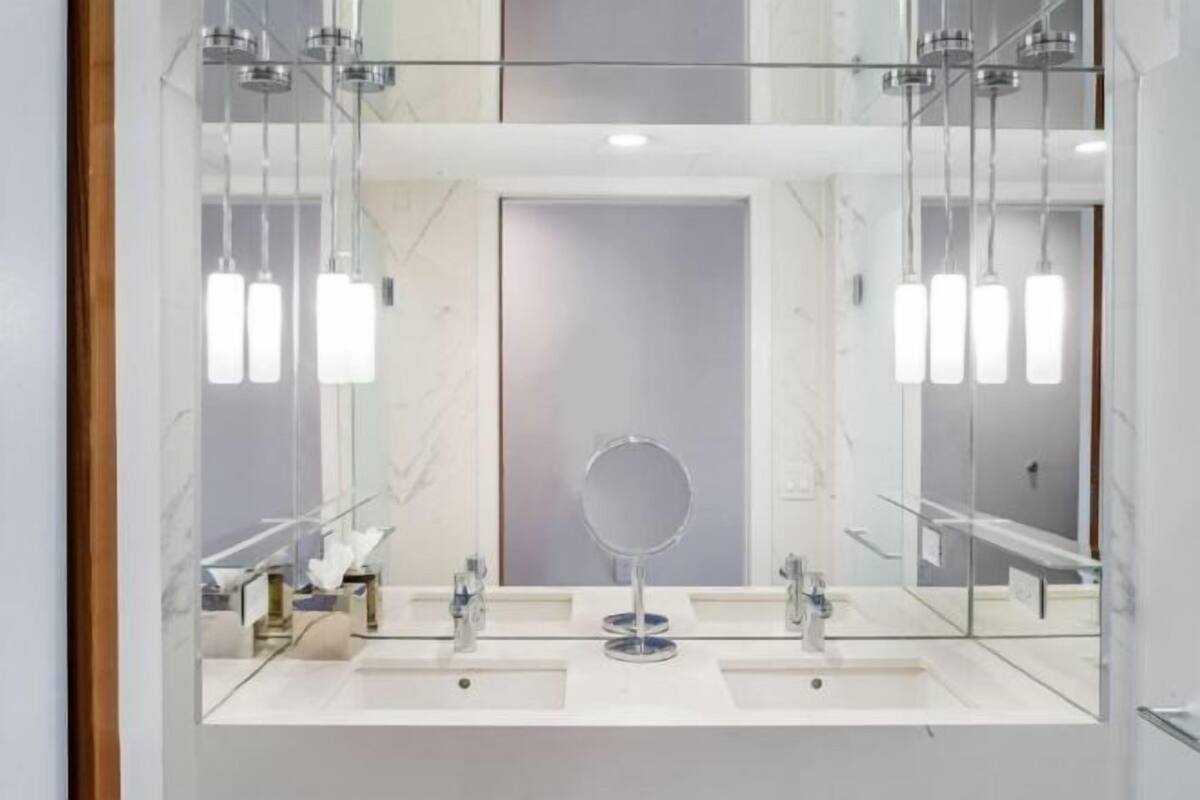 Uma casa de banho moderna com cabine de vidro, duas pias, espelho redondo e luzes pendentes.