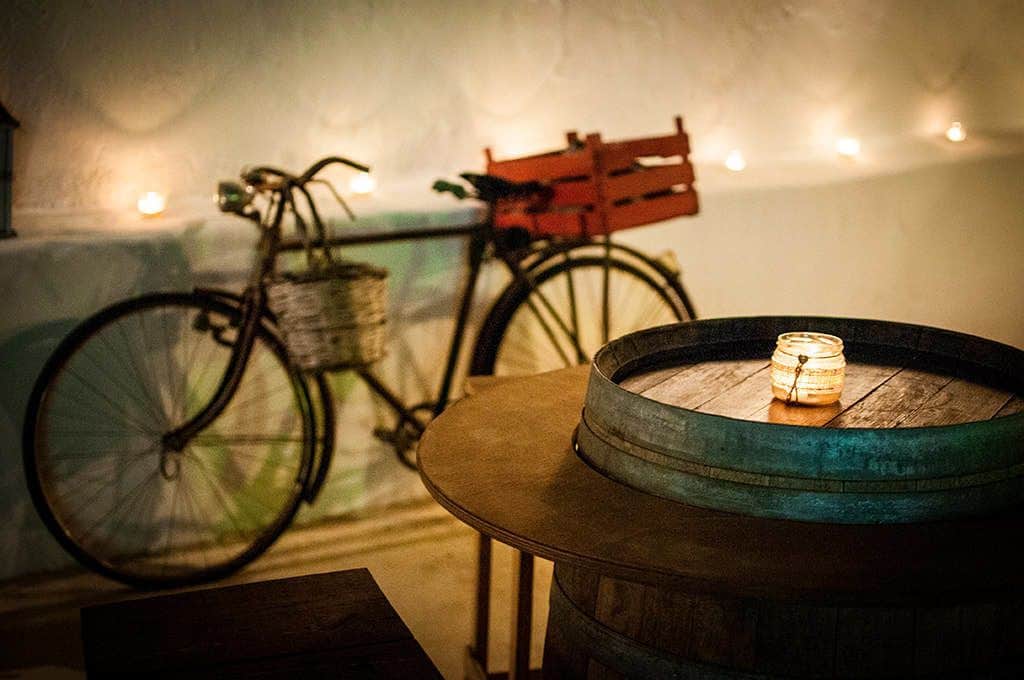 Um interior rústico com uma mesa de madeira, um barril de metal, uma bicicleta e uma caixa vermelha.