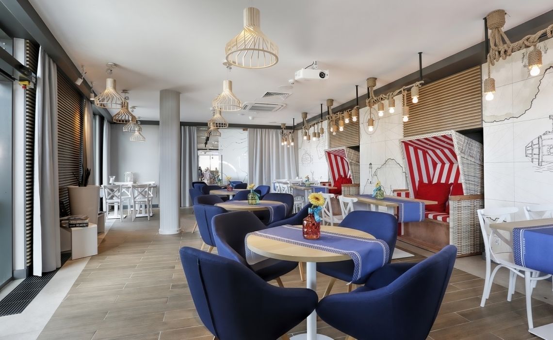 Interior de restaurante moderno com mesas redondas, cadeiras azuis e luminárias pendentes.