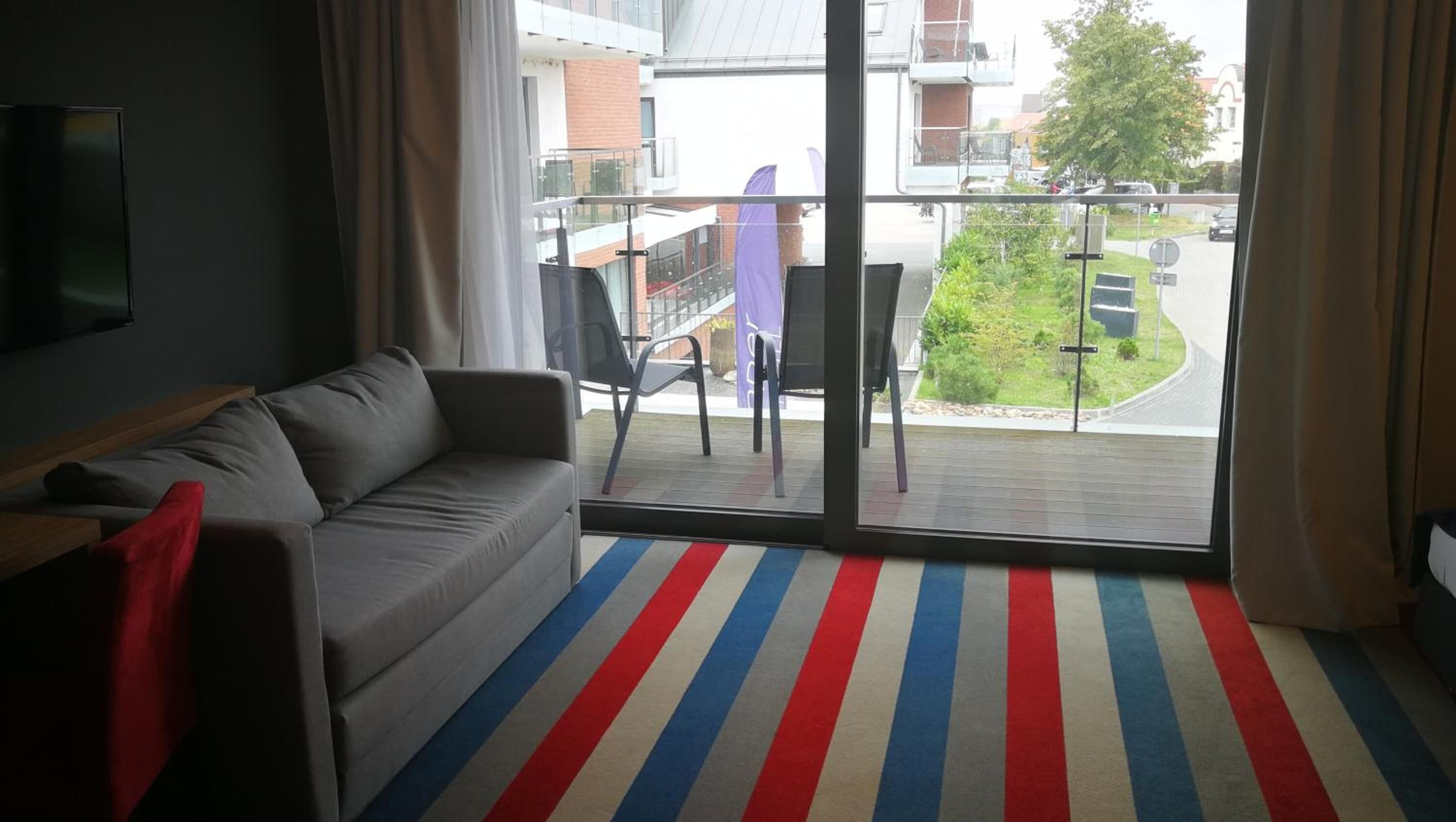 Quarto de hotel moderno com um sofá cinza, um tapete listrado e uma varanda com vista para a rua e edifícios.