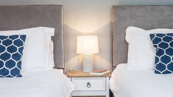 Uma cama de casal com roupa de cama branca e almofadas estampadas em azul, uma mesa de cabeceira com abajur e uma gaveta pequena.