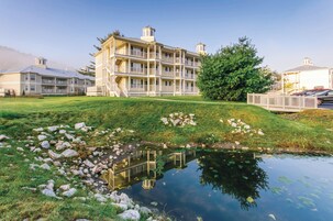 Um edifício de vários andares com uma varanda, um lago com pedras e uma árvore.