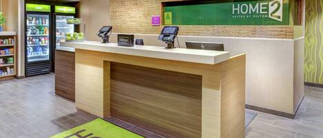 Uma área de recepção de hotel com balcão de atendimento, um quiosque digital e uma placa indicando 'HOME 2 Suites by Hilton'. Há duas geladeiras identificadas como 'fria' e 'bem fria', com bebidas e lanches. Um tapete verde com 'HOME 2' está em frente ao balcão.