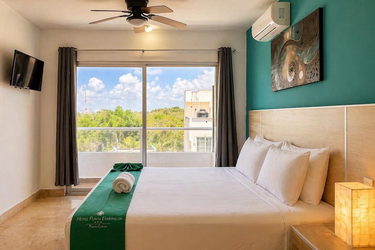 Quarto de hotel com cama, toalha verde, ventoinha no teto, televisão e vista para edifícios e árvores.