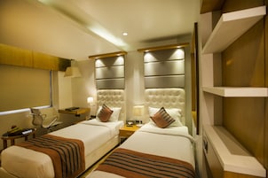 Quarto de hotel com duas camas, uma escrivaninha, uma cadeira, um abajur e uma prateleira.