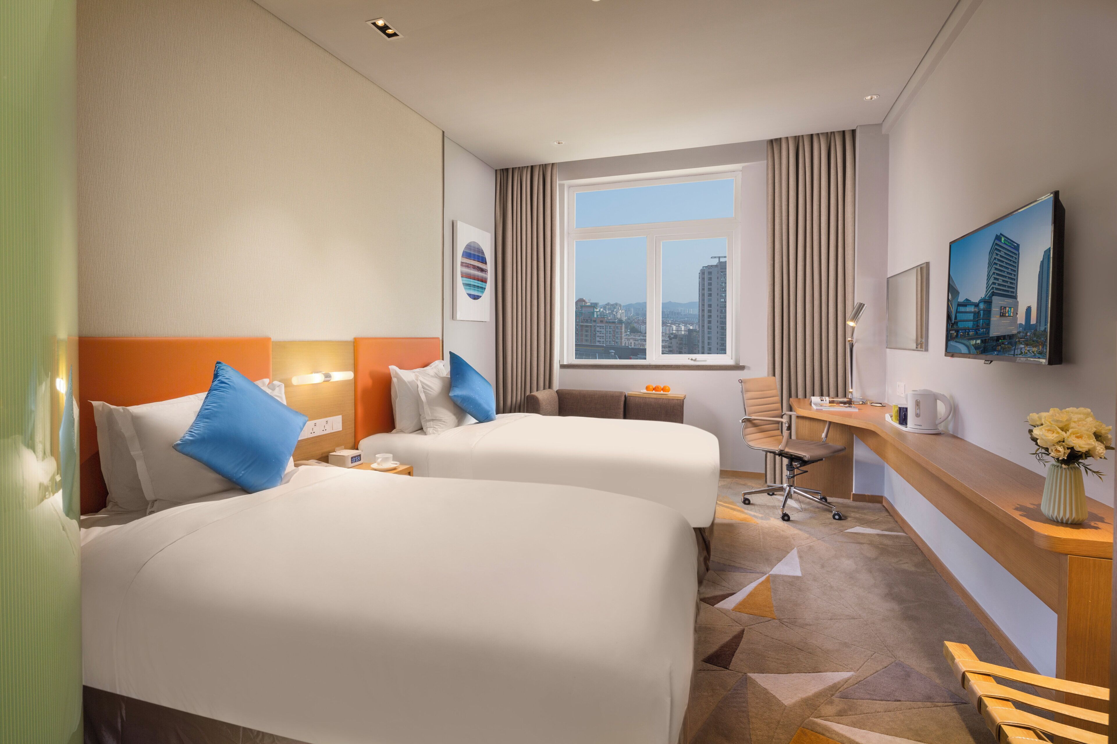 Quarto de hotel com duas camas, uma escrivaninha, uma cadeira, janela com vista para a cidade e um quadro na parede.
