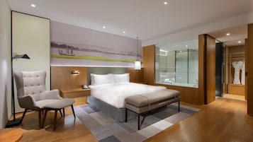 Quarto de hotel moderno com uma cama grande, área de estar com uma poltrona, mesa de cabeceira e luminária de parede.