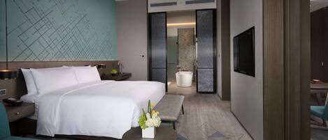 Quarto de hotel moderno com uma cama grande, uma escrivaninha, uma televisão e um banheiro visível através de uma porta de vidro.