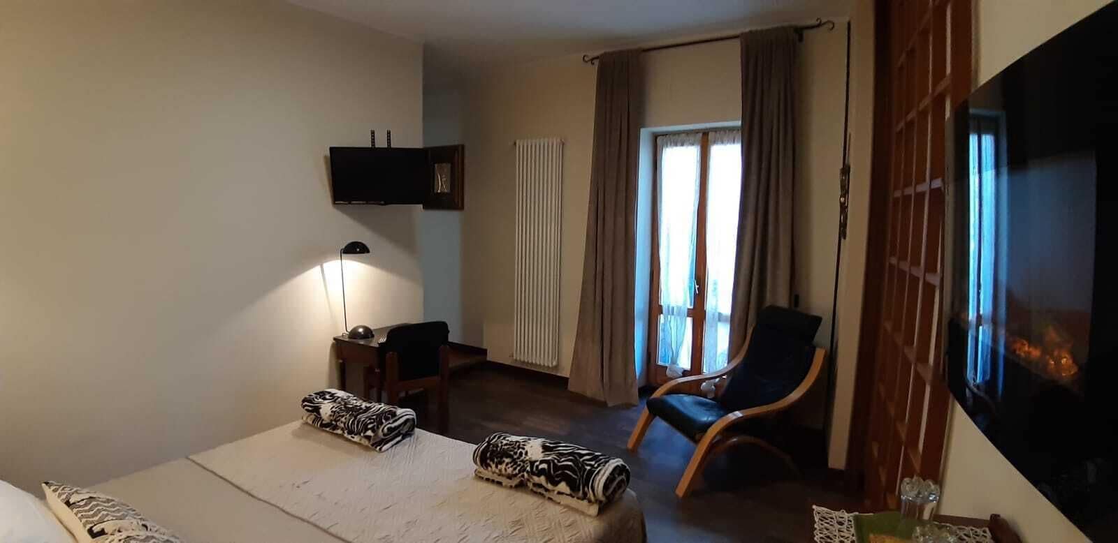 Quarto de hotel com cama, cadeira, televisão, luminária e janela com cortinas.