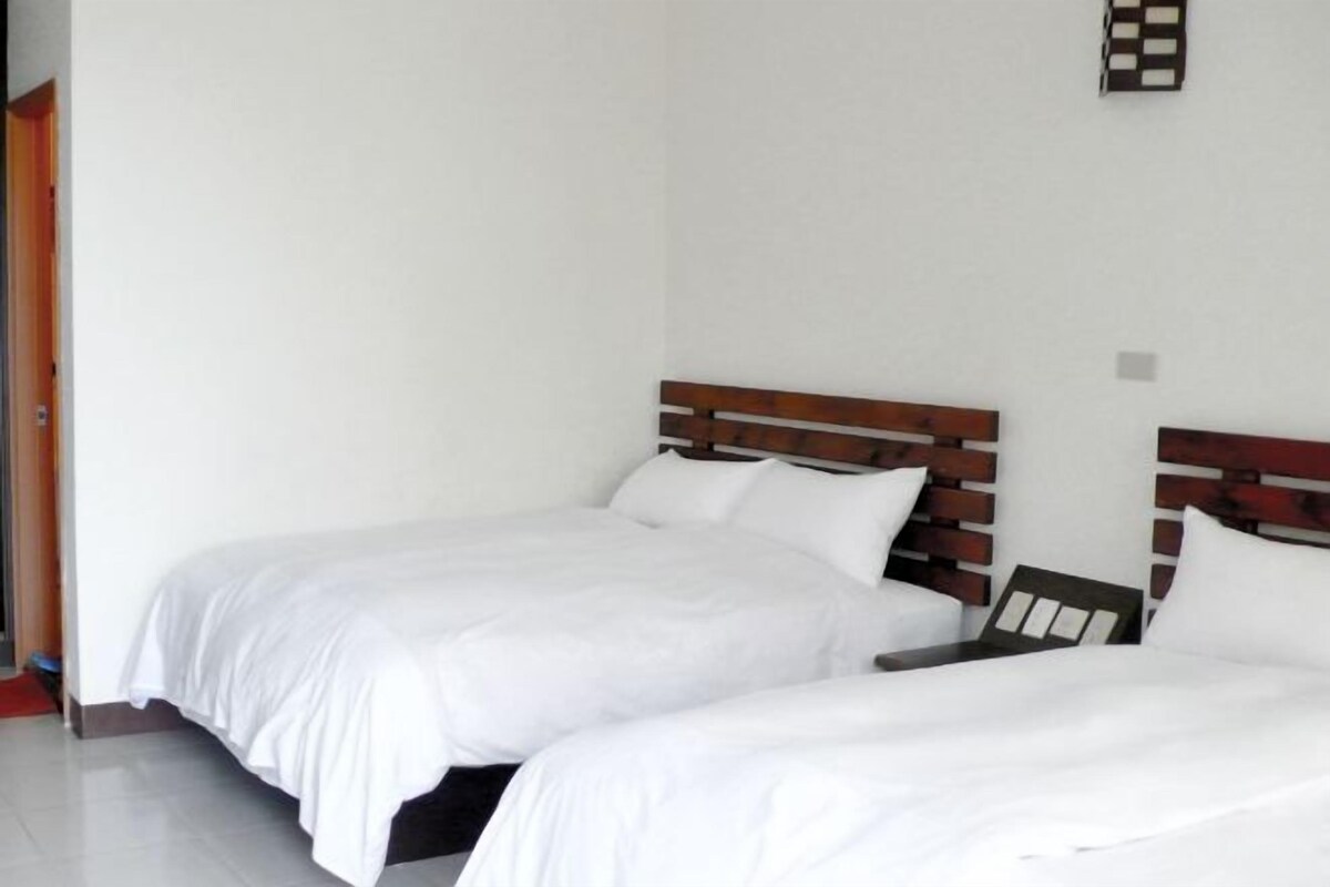 Quarto de hotel com duas camas de solteiro, cabeceiras de madeira, roupa de cama branca e uma pequena mesa de cabeceira.