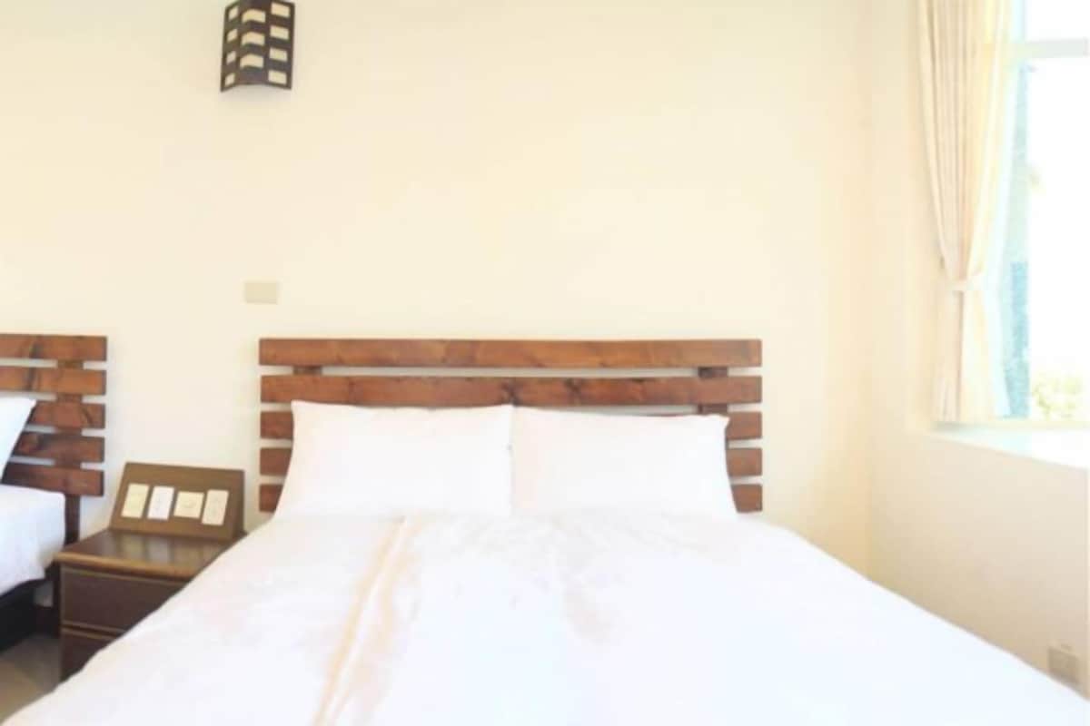 Quarto de hotel com cabeceira de madeira, cama com roupa de cama branca, mesa de cabeceira com abajur e janela com cortinas.