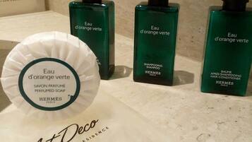 Três frascos de shampoo e condicionador verde, um sabonete branco em barra e um saco de papel branco com os dizeres "Eau d'orange verte" e "Hermès Paris" sobre uma superfície de mármore.