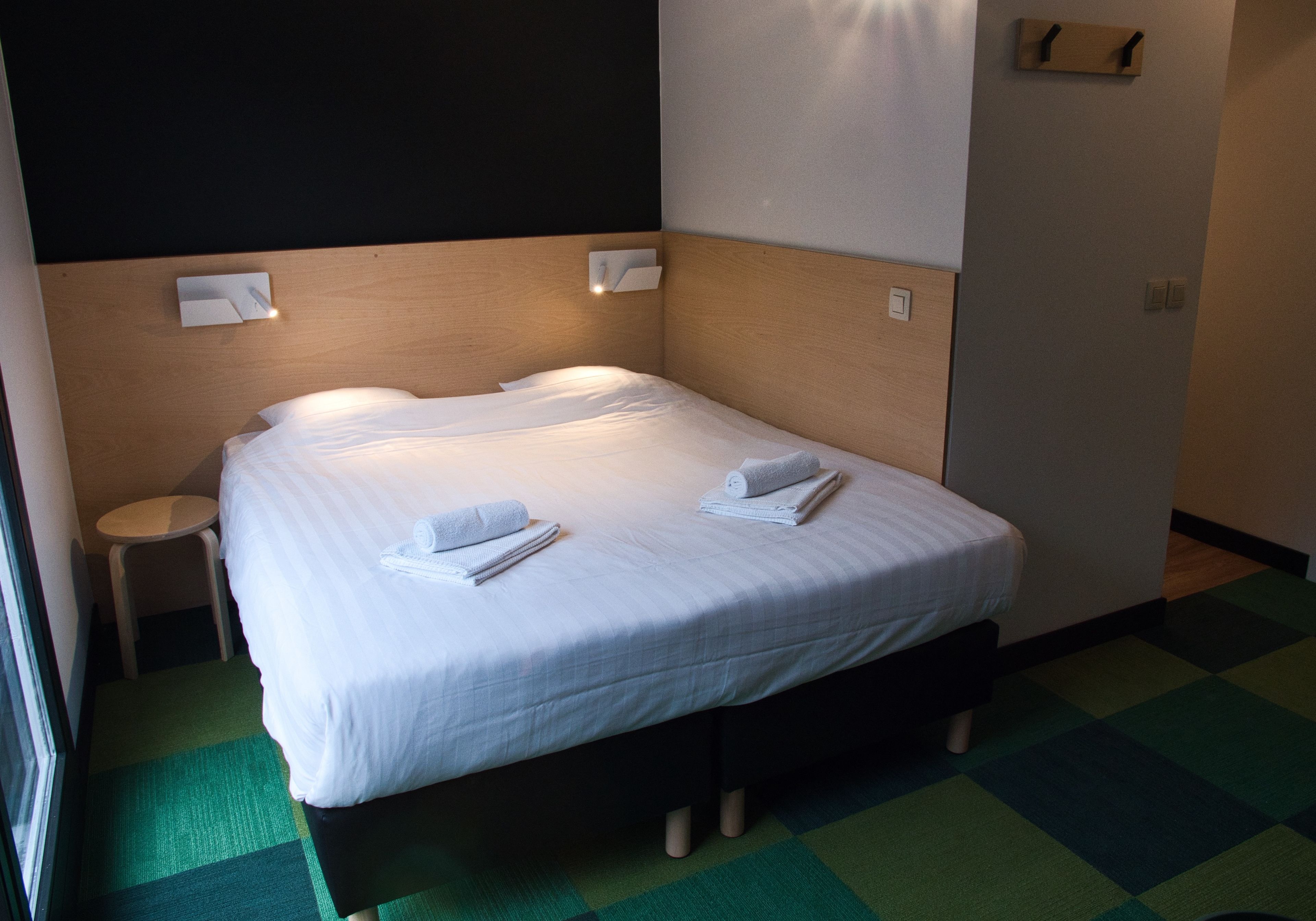 Uma cama de casal bem arrumada com lençóis e toalhas brancas, uma pequena mesa de cabeceira e um chão quadriculado verde e azul.