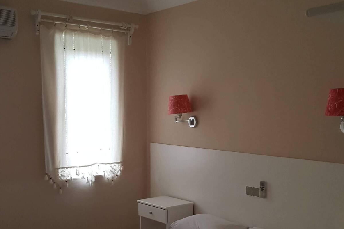Quarto com cama, mesa de cabeceira, luminária na parede e janela com cortinas.