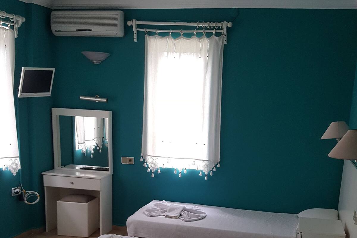 Quarto com duas camas, uma escrivaninha, uma cadeira, uma televisão e uma janela com cortinas.