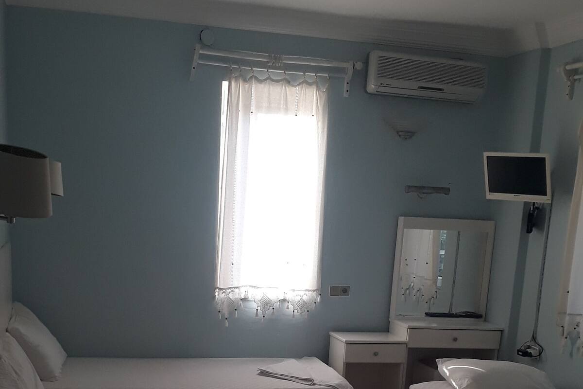 Quarto de hotel com duas camas, janela com cortinas leves, televisão de tela plana, mesa de cabeceira pequena e unidade de ar condicionado.