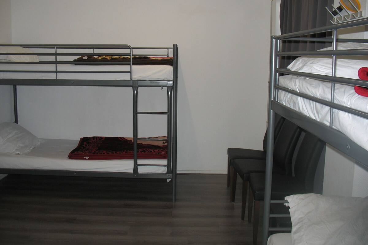Quarto com beliche, escrivaninha e cadeira, uma raquete de ténis sobre a escrivaninha e uma janela com cortinas.