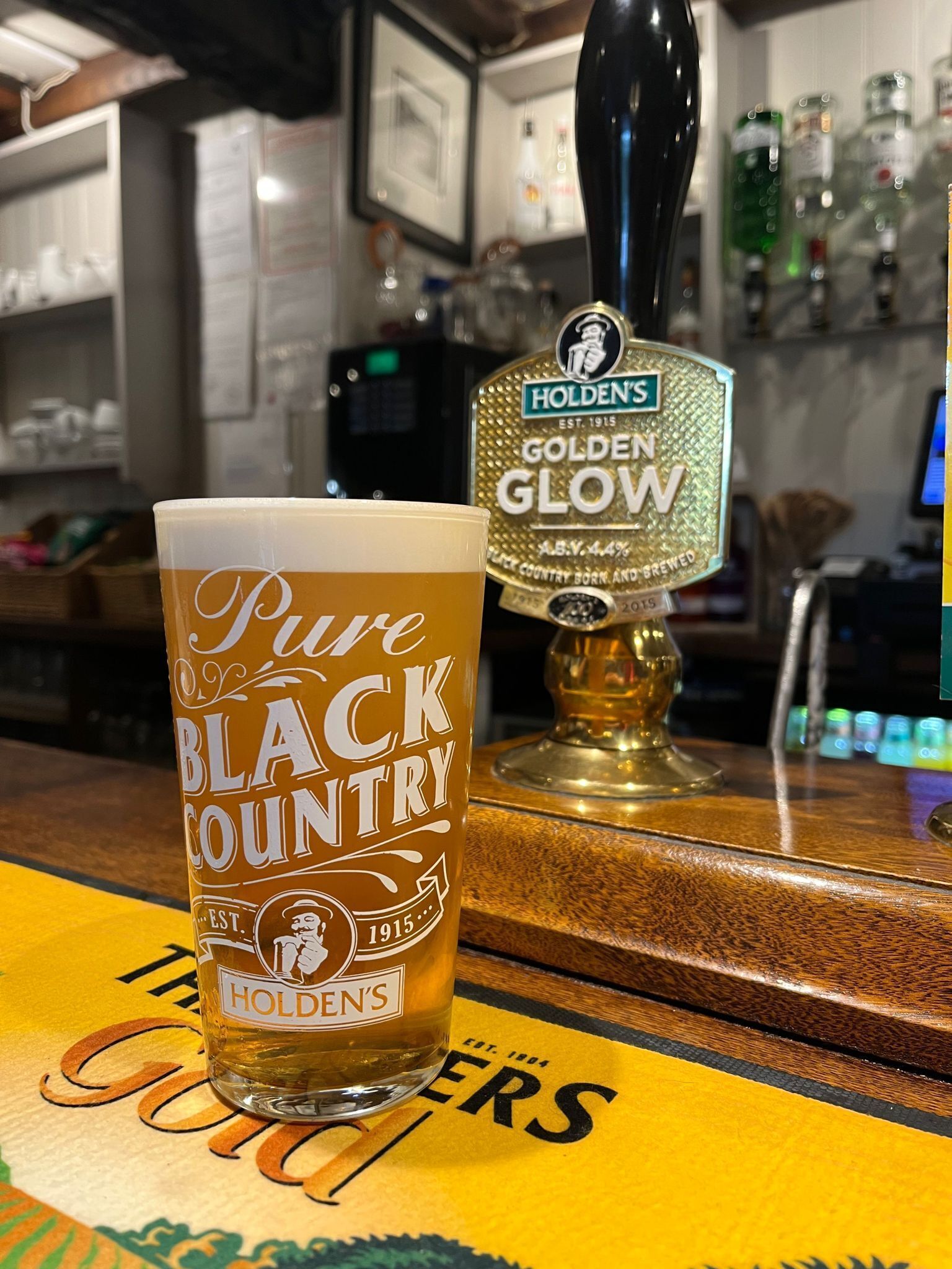 Um copo de cerveja com a marcação 'Pure Black Country' e uma torneira identificada como 'Holden's Golden Glow'.