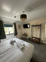 Quarto de hotel com cama, televisão, prateleira e janela com cortinas.