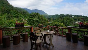 Terraço com mobiliário de madeira e plantas em vasos, com vista para uma paisagem verde exuberante e montanhas ao longe.
