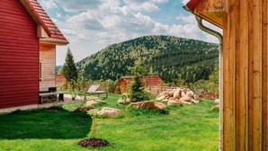 Uma cabana de madeira com telhado vermelho, um gramado e uma montanha arborizada ao fundo.