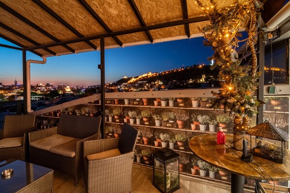 Terraço no telhado com móveis de vime, uma mesa redonda e diversos vasos de plantas.