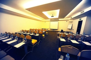 Uma sala de conferências com várias mesas e cadeiras, uma tela de projetor e um quadro branco.