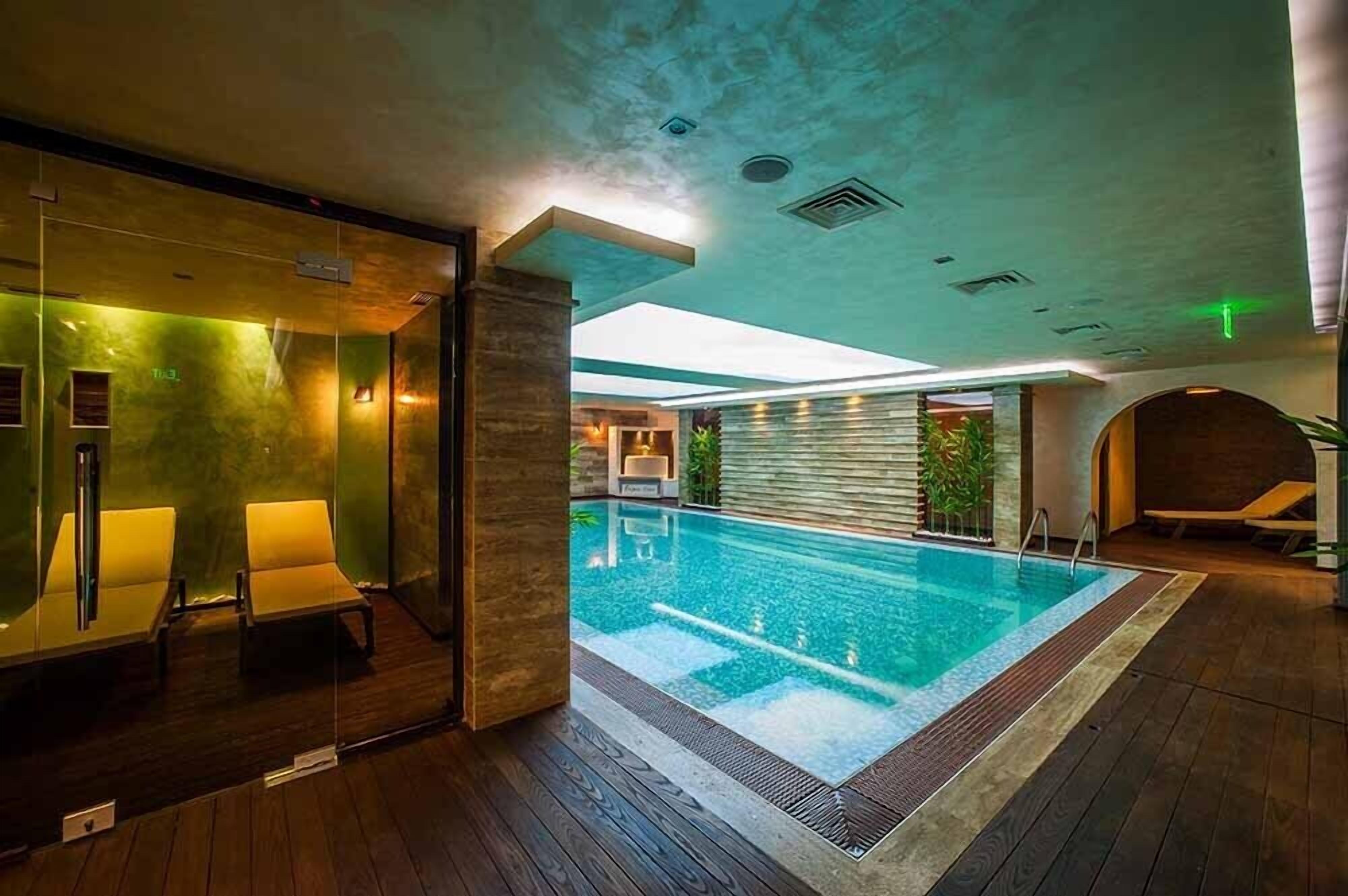 Uma área de piscina interior moderna com estrutura de vidro, piso de madeira e espreguiçadeiras.