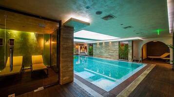 Uma área de piscina interior moderna com estrutura de vidro, piso de madeira e espreguiçadeiras.