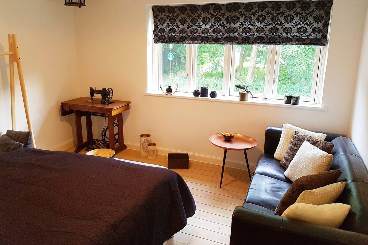 Um quarto aconchegante com uma cama, uma mesa de madeira com uma máquina de costura, uma mesinha com uma tigela, um sofá com almofadas e uma janela com vista para o verde.
