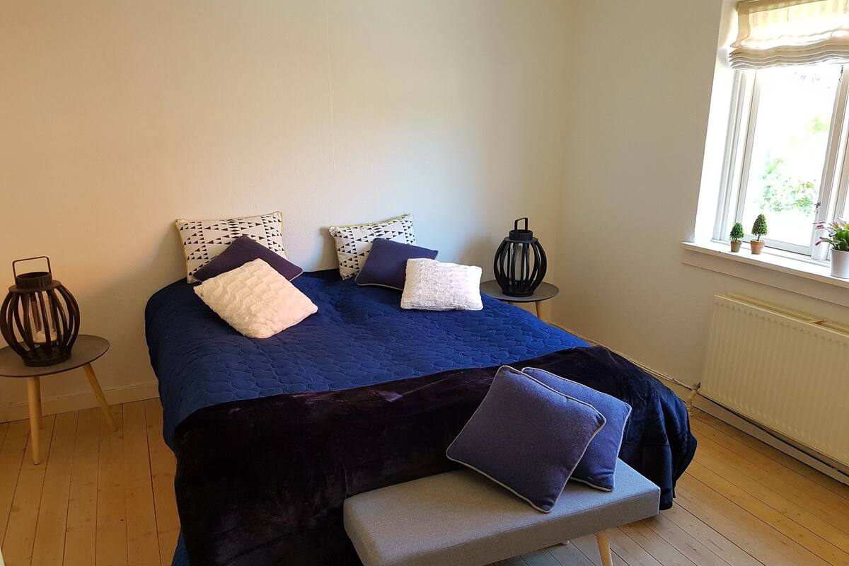 Quarto com cama, um banco, uma pequena mesa com um candeeiro e uma janela com plantas.