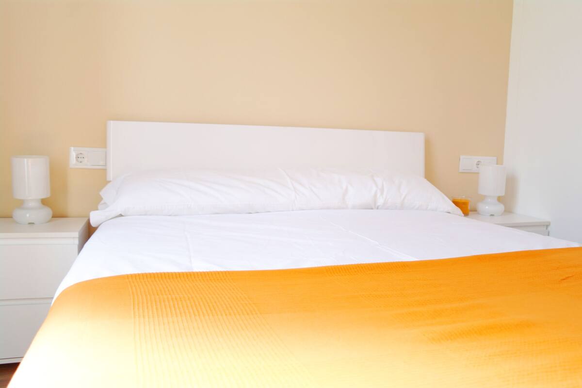 Cama com roupa de cama branca e cabeceira branca, com dois mesinhas de cabeceira brancas com luminárias.