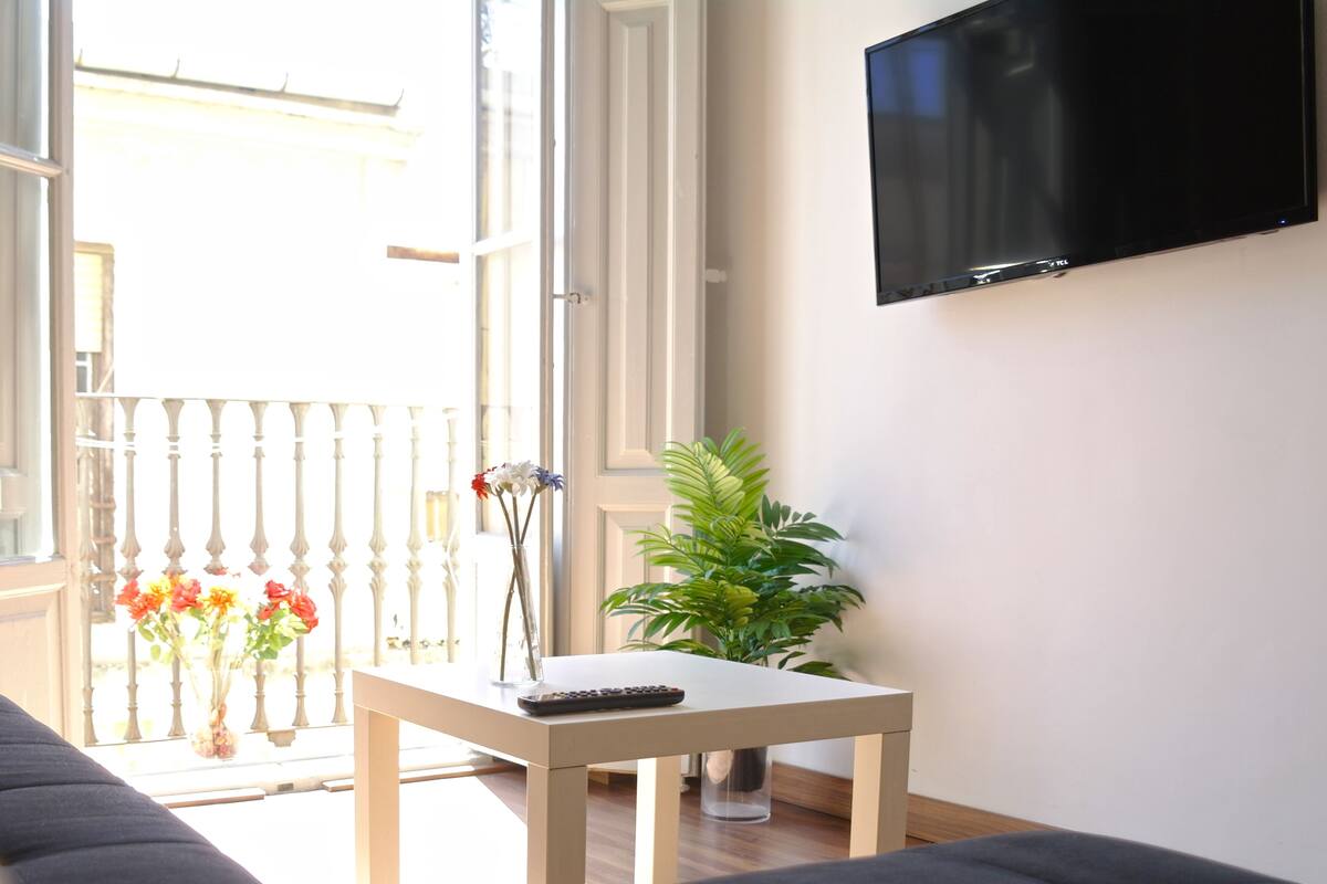 Uma sala de estar com televisão de tela plana na parede, uma mesinha com um vaso de flores e uma varanda com guarda-corpo e flores.