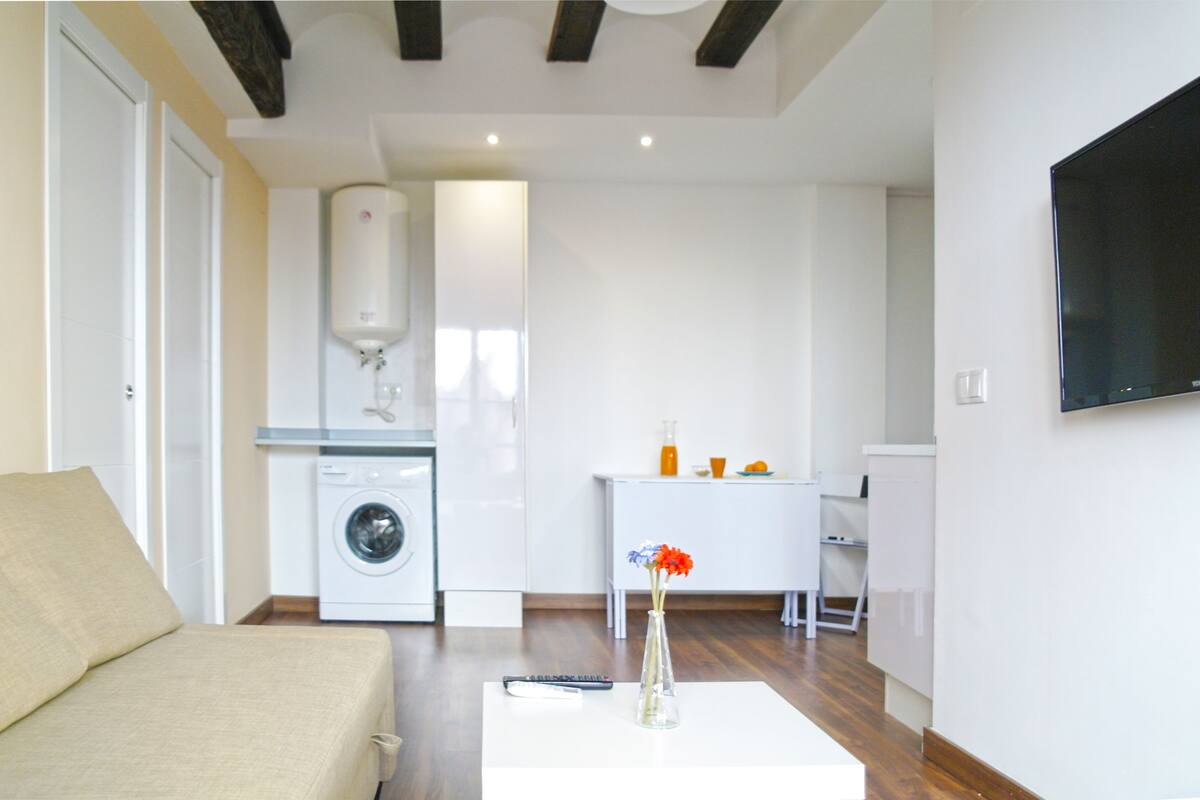 Uma sala de estar moderna com sofá, mesa de centro com um vaso, máquina de lavar, mesa de jantar e televisão na parede.