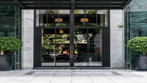 Entrada de um edifício moderno com portas de vidro, galeria coberta e plantas em vasos.