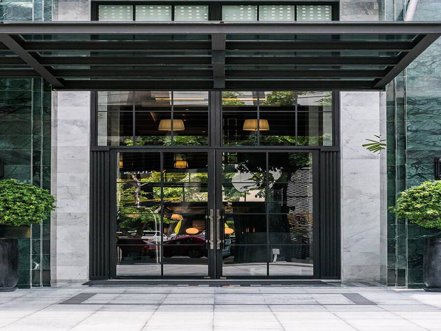 Entrada de um edifício moderno com portas de vidro, galeria coberta e plantas em vasos.