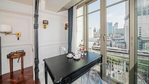 Quarto com uma mesa preta posta para chá, vista da cidade através de janelas amplas e uma parede branca com luminária dourada.