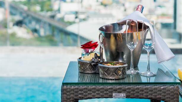 Mesa com balde de champanhe, copos e arranjo de flores ao lado de uma piscina com vista para a cidade.