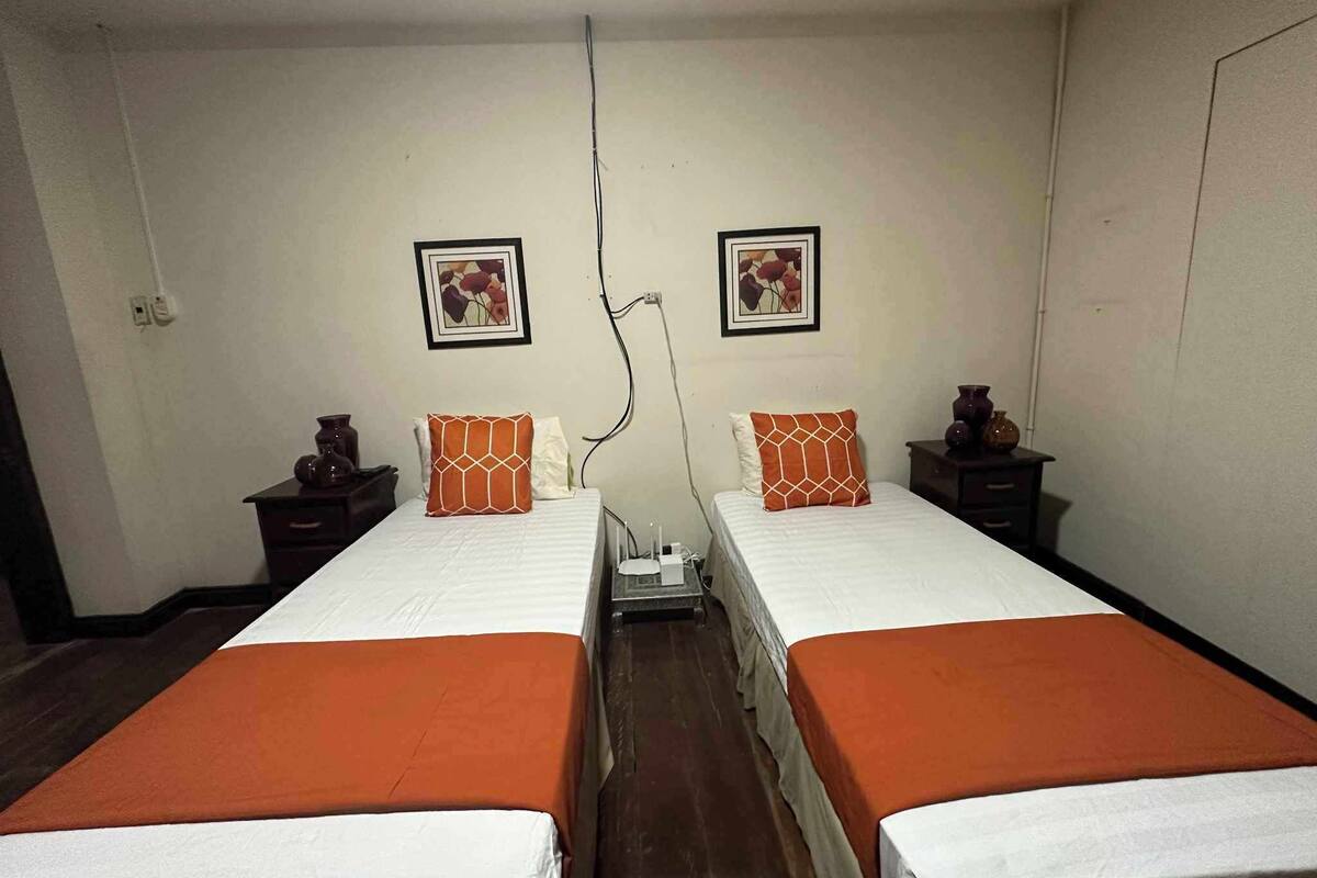 Quarto com duas camas, cada uma com colcha laranja e roupa de cama branca. Há duas mesas de cabeceira com objetos decorativos e duas fotos emolduradas na parede.