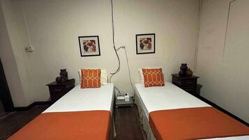 Quarto com duas camas, cada uma com colcha laranja e roupa de cama branca. Há duas mesas de cabeceira com objetos decorativos e duas fotos emolduradas na parede.