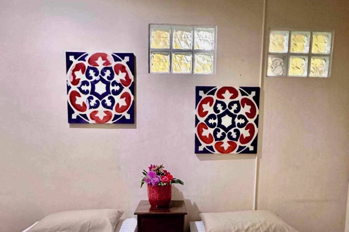 Quarto com duas camas, uma pequena mesa de madeira com um vaso de flores e duas janelas embutidas na parede.