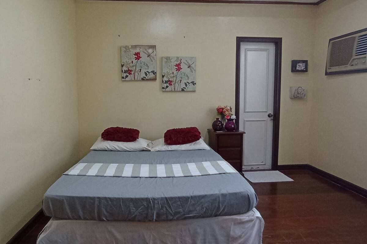 Um quarto com uma cama, duas pinturas nas paredes, uma porta e um aparelho de ar condicionado.
