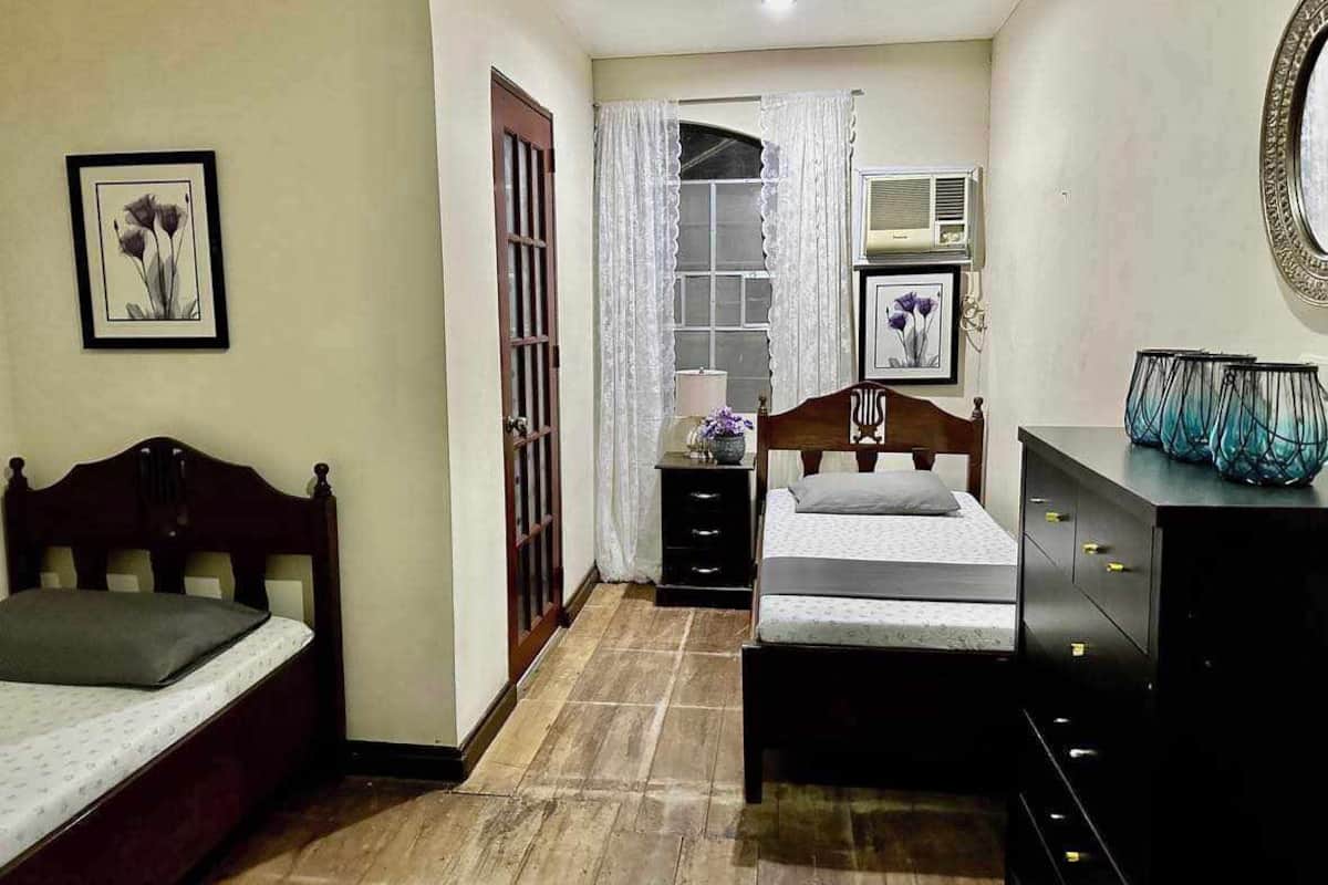 Quarto com duas camas, uma cômoda, um espelho e uma janela com cortinas.