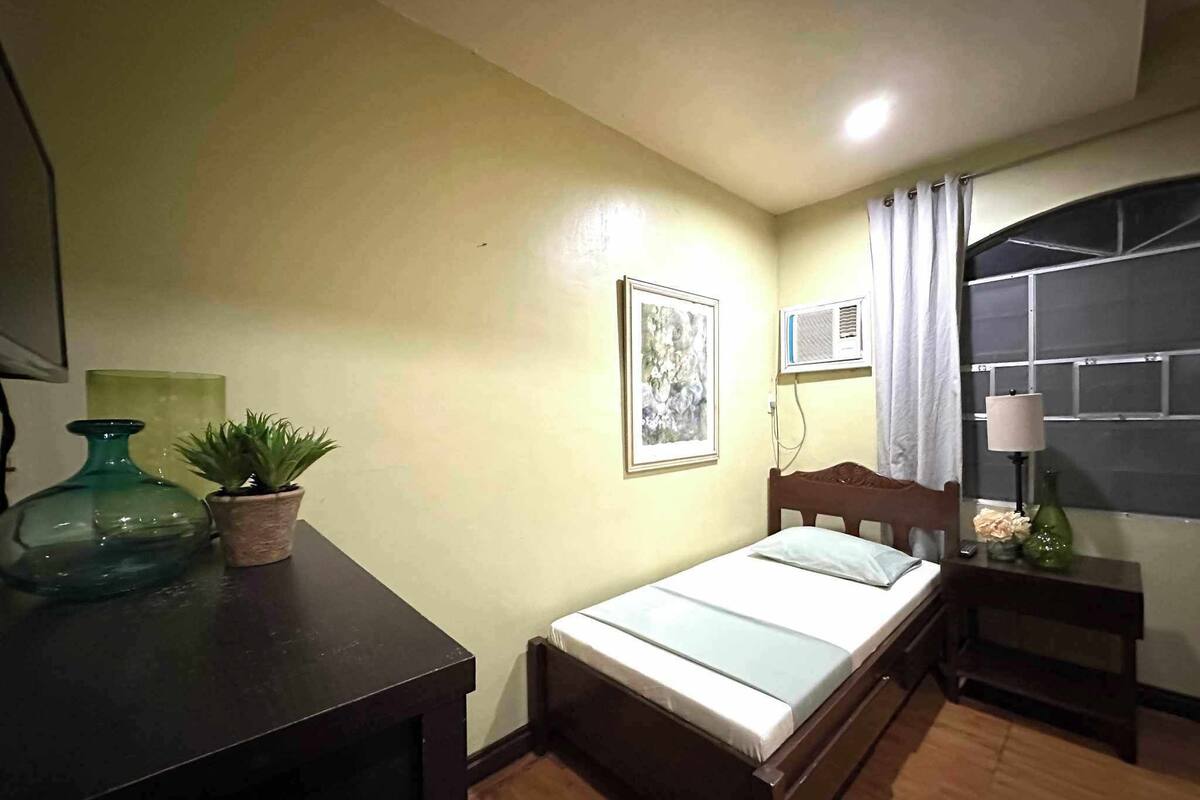 Quarto pequeno e bem arrumado com uma cama de solteiro, mesa de cabeceira, luminária, um vaso de planta, um quadro na parede e um aparelho de ar condicionado.