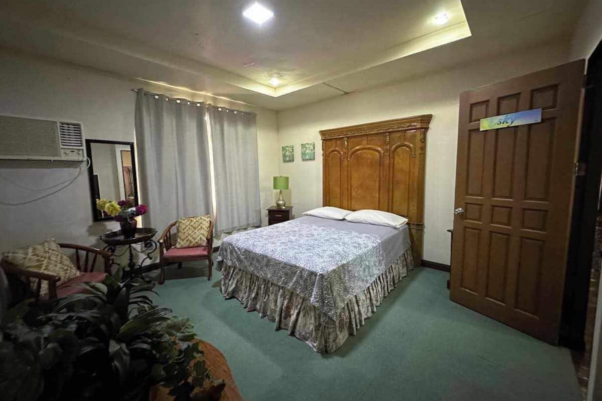 Quarto de hotel com cama, escrivaninha, cadeira, planta em vaso, mesa de cabeceira e guarda-roupa de madeira.