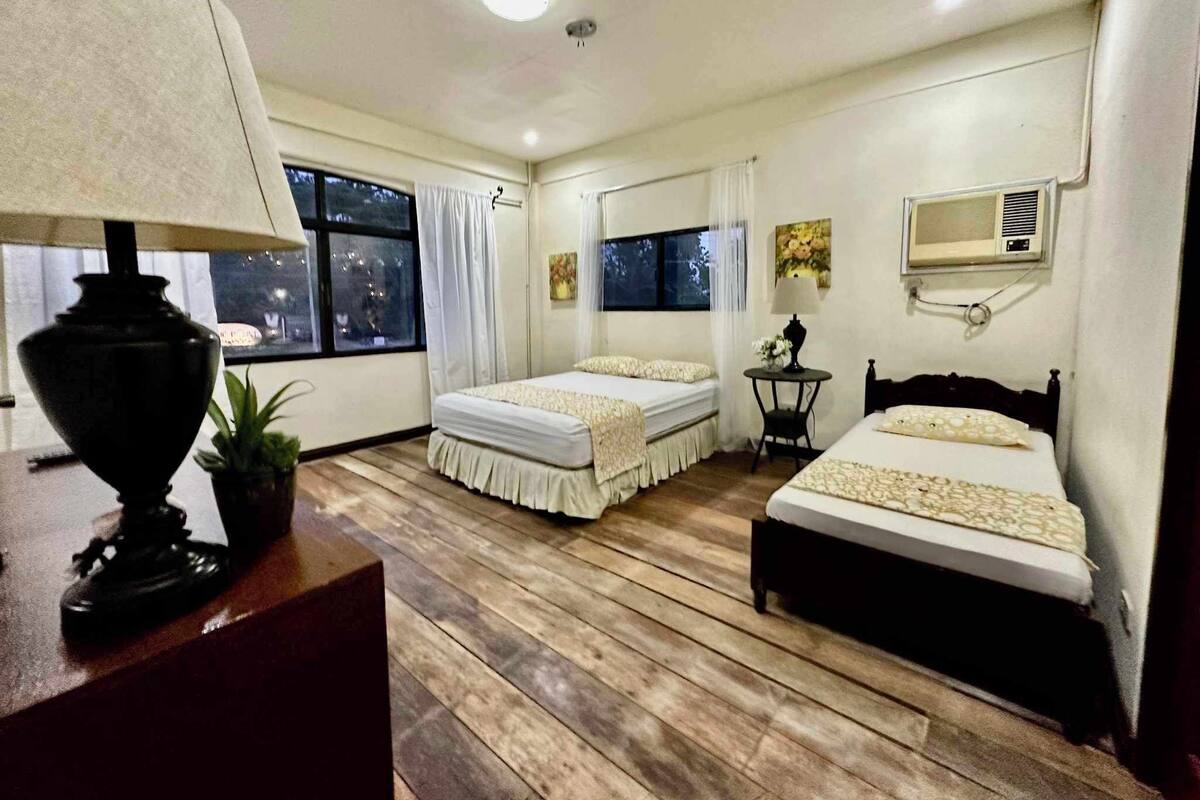 Quarto de hotel com duas camas, uma janela grande, uma luminária de teto e uma mesa pequena com abajur.
