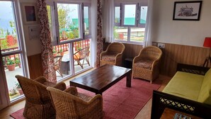 Quarto com mobiliário de vime, uma mesa de madeira e vista para um jardim através de grandes janelas.